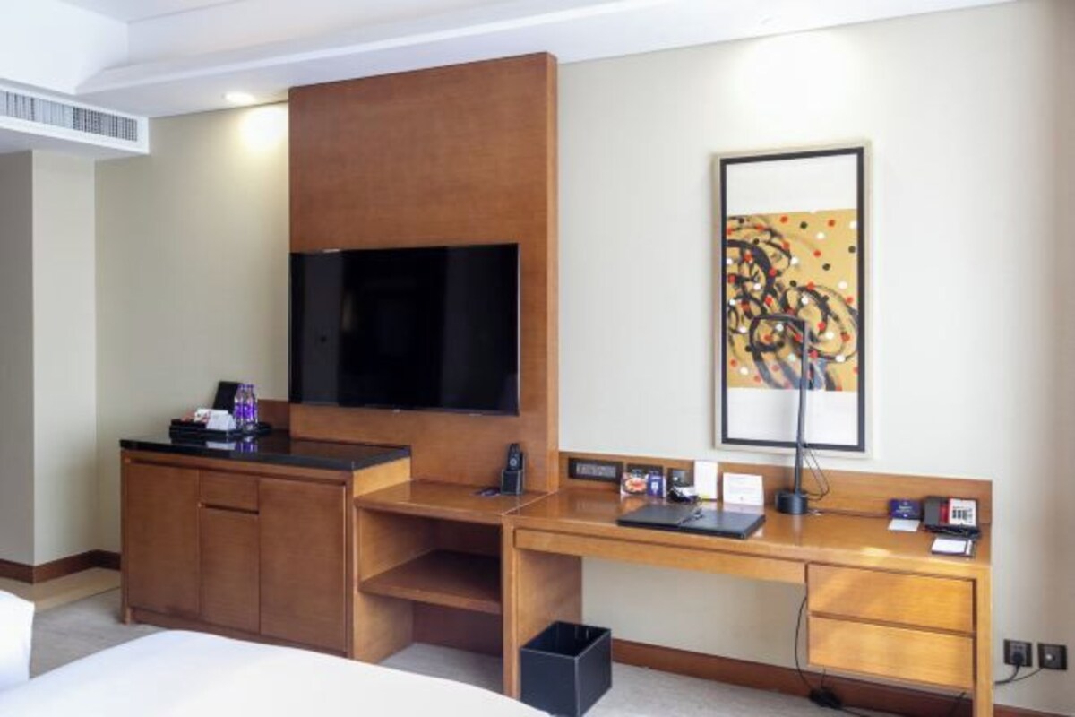 Quarto de hotel com cama, escrivaninha de madeira, televisão de tela plana, pintura abstrata emoldurada e uma lixeira pequena.