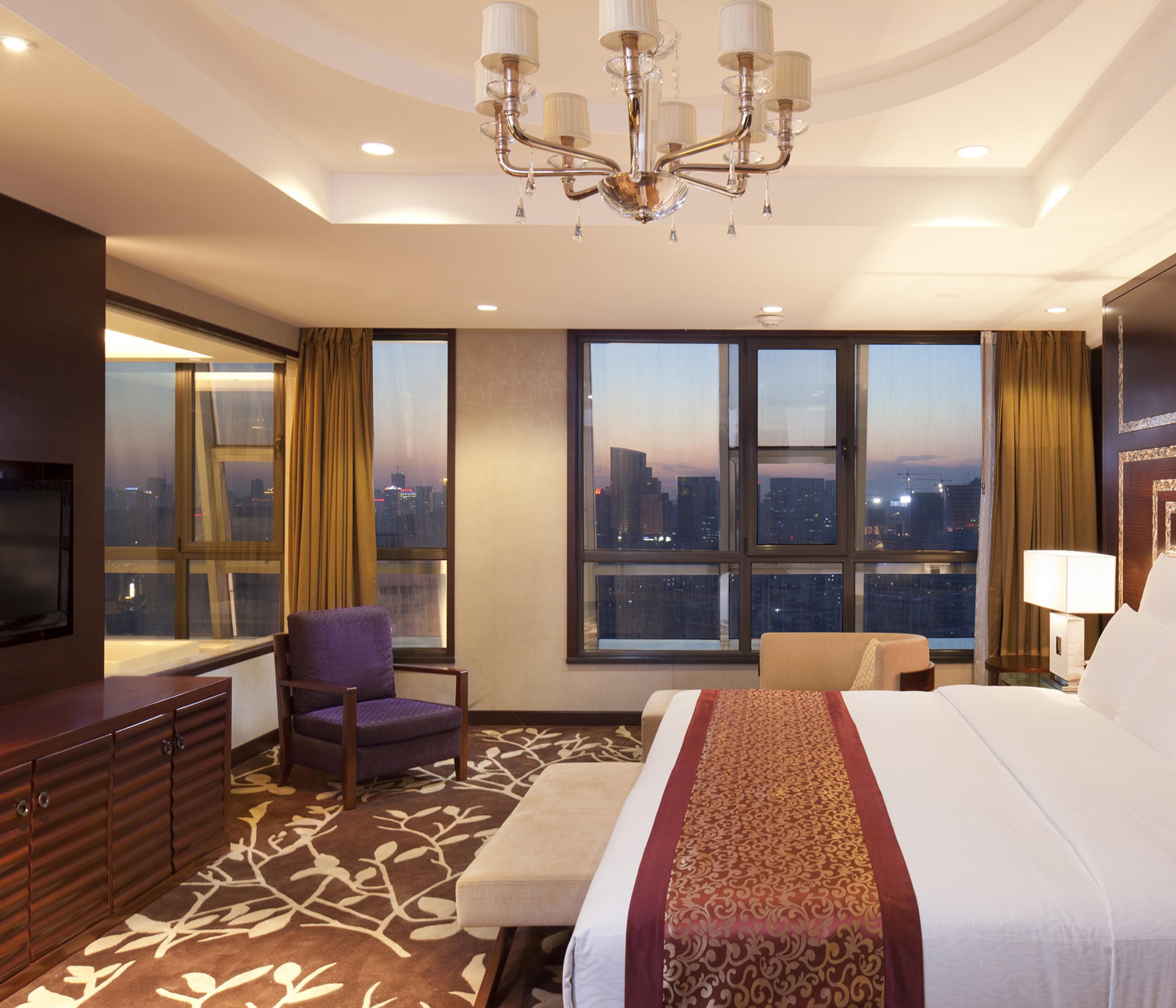 Quarto de hotel com uma cama grande, televisão, cadeira, escrivaninha e vista para a paisagem urbana pelas janelas.