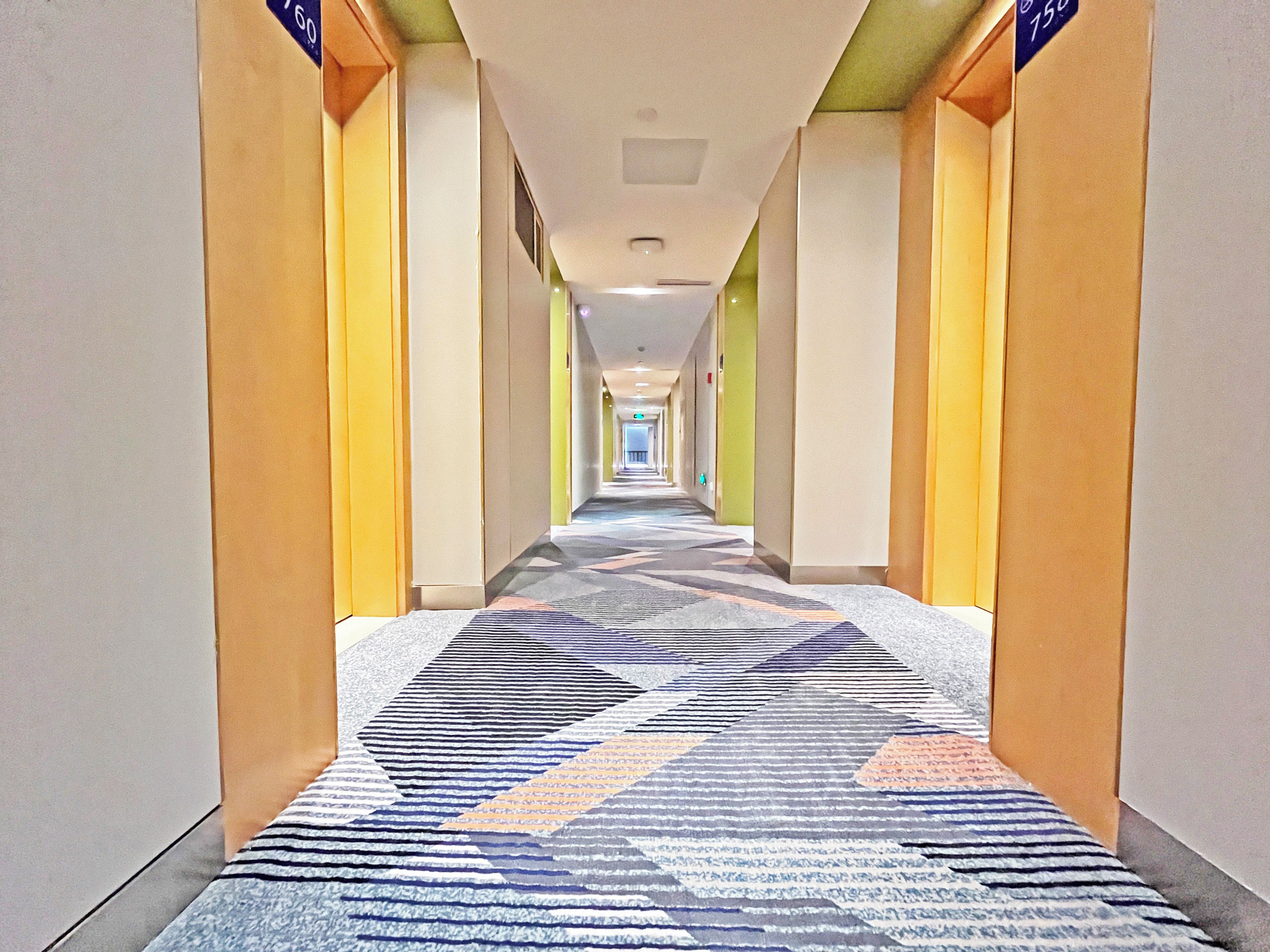 Corredor de hotel com carpete estampado, portas de ambos os lados e uma placa indicando os números dos quartos.