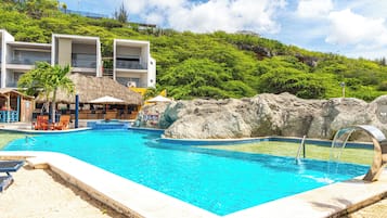 Piscina com uma formação rochosa, espreguiçadeiras e um edifício com varandas.