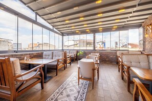 Restaurante no terraço com mobiliário de madeira, grandes janelas e vista para a cidade.
