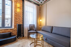 Um quarto com parede de tijolos, sofá, mesa de vidro, cadeira, radiador e janela com cortinas.