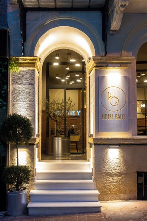 Entrada de hotel com uma placa indicando 'HOTEL BALO', uma escadaria que leva para dentro e um vaso de planta à esquerda.