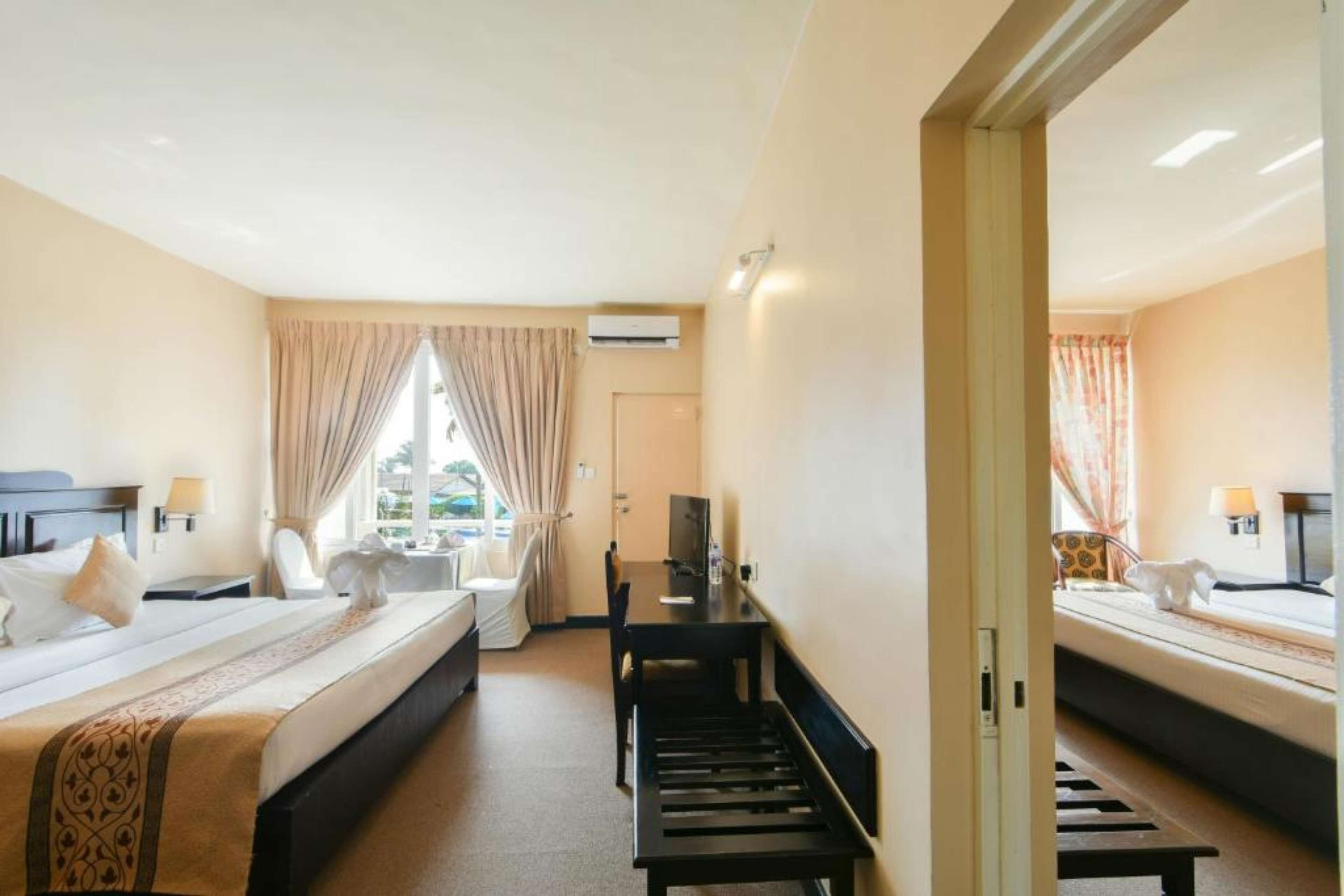 Quarto de hotel com duas camas, uma escrivaninha e uma cadeira. Há uma janela com cortinas, um aparelho de ar condicionado e vista para o exterior.