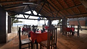 Uma área de refeições ao ar livre com mesas e cadeiras, vista para a praia e estrutura com telhado de palha.