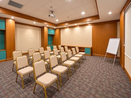 Sala de conferências com fileiras de cadeiras, um quadro branco e piso coberto de carpete.
