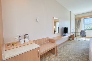 Quarto de hotel com um banco de madeira, televisão, escrivaninha e janela com vista para árvores.