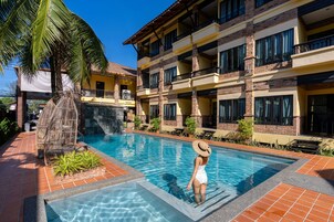 Piscina com uma pessoa na beira, cercada por um edifício com várias varandas e uma palmeira.