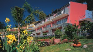 Um hotel com vários andares, varandas, um jardim com flores e plantas, e um céu azul claro.