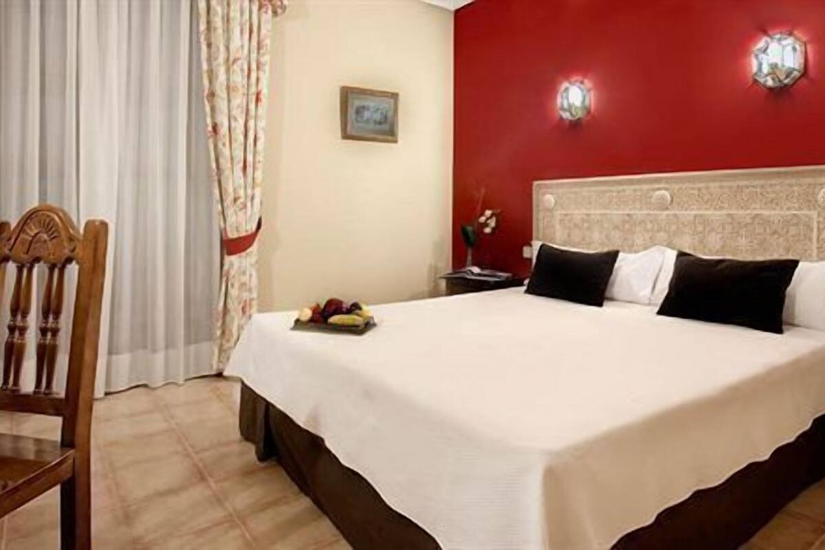 Quarto de hotel com uma cama grande, uma cadeira de madeira, uma mesa de cabeceira e uma parede vermelha.
