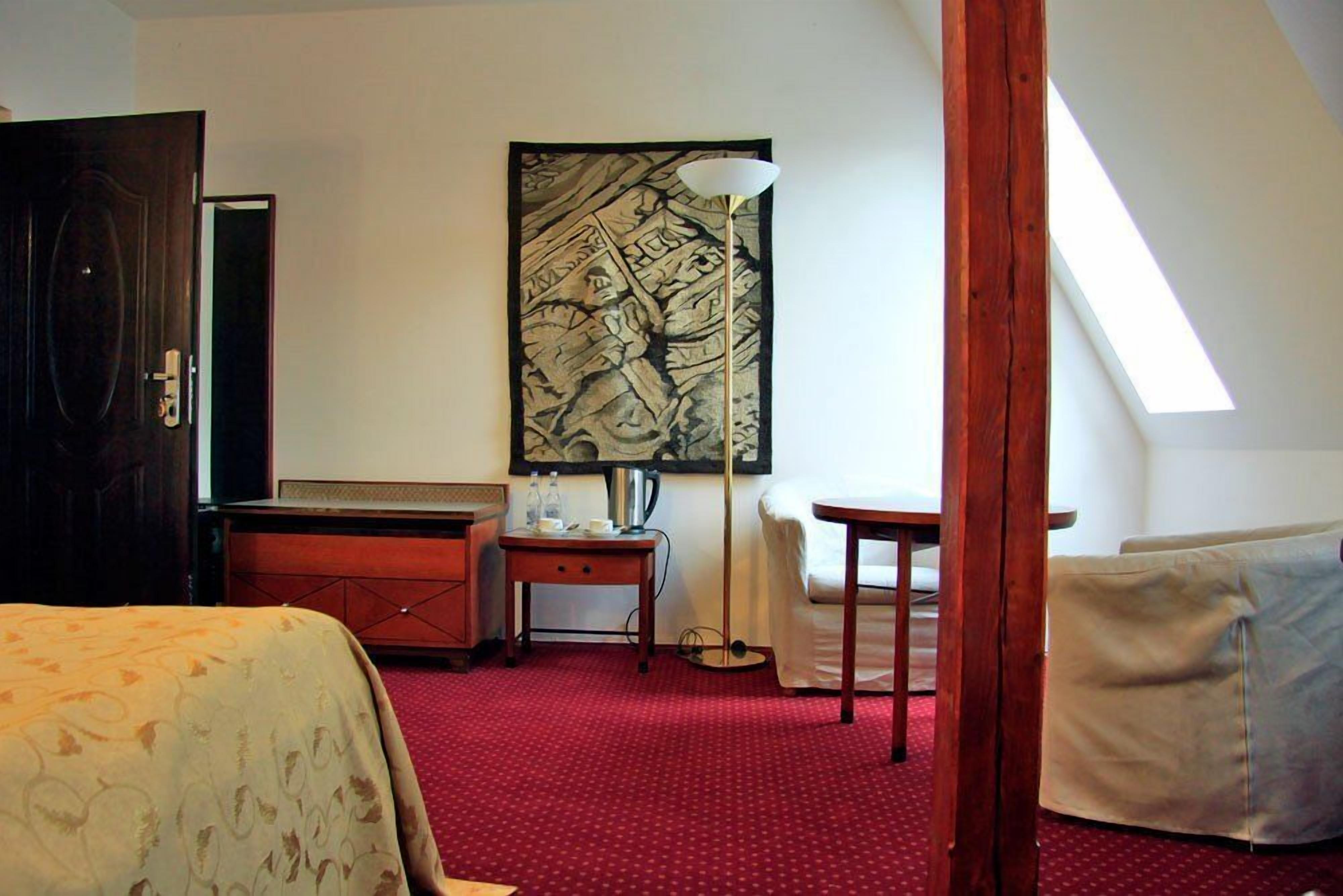 Quarto de hotel com carpete vermelho, cama com colcha estampada, escrivaninha com luminária, pintura na parede e janela com luz natural.