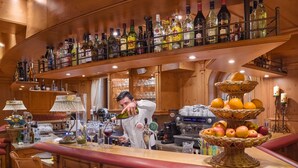 Uma área de bar com um bartender servindo uma bebida, prateleiras com diversos frascos e uma exposição de frutas.