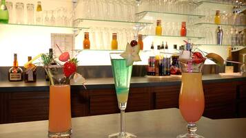 Três cocktails sobre um balcão de bar, com diversos enfeites, e ao fundo, estantes com garrafas e copos.
