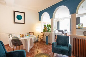 Uma sala de jantar com uma mesa posta para dois, uma planta em vaso e vista através de um arco para outro ambiente.