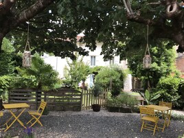 Um pátio com jardim, com cadeiras e mesas amarelas, cercado por árvores e um portão de madeira.