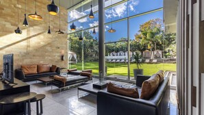 Uma área de estar moderna com sofás de couro, uma televisão de tela plana e luminárias pendentes. Janelas amplas oferecem vista para um gramado bem cuidado e área de estar externa.