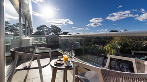 Varanda com mesa posta para o café da manhã, incluindo torradas, suco e uma xícara, com vista para uma paisagem cênica com árvores e céu claro.