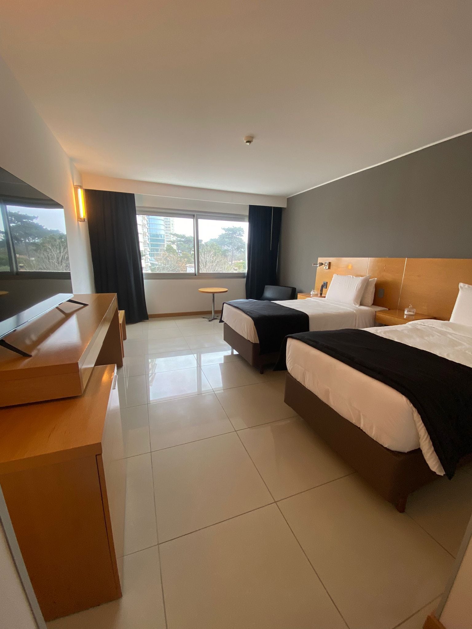 Quarto de hotel com duas camas, uma escrivaninha, uma cadeira, uma mesinha e uma janela com cortinas.