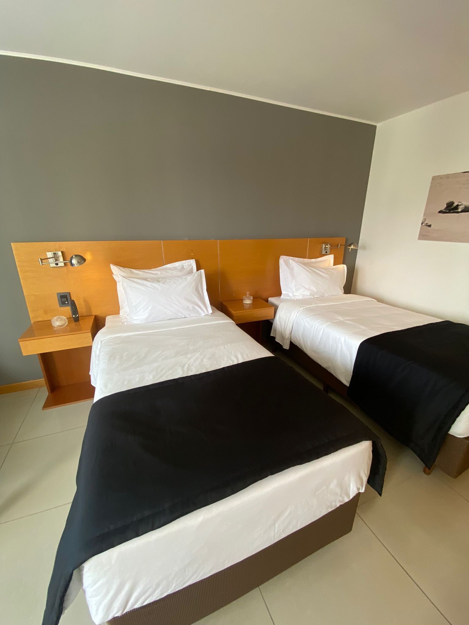 Quarto de hotel com duas camas, cabeceiras de madeira, mesas de cabeceira e uma luminária fixada na parede.