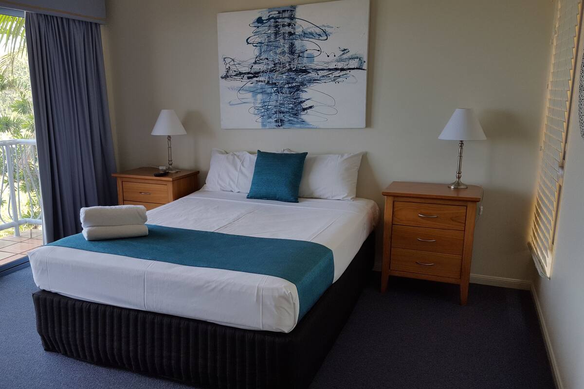 Quarto de hotel com uma cama grande, duas mesas de cabeceira com luminárias, uma janela com cortinas e um quadro na parede.