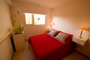 Quarto com colcha vermelha, duas almofadas, mesa de cabeceira com abajur e janela com persianas.