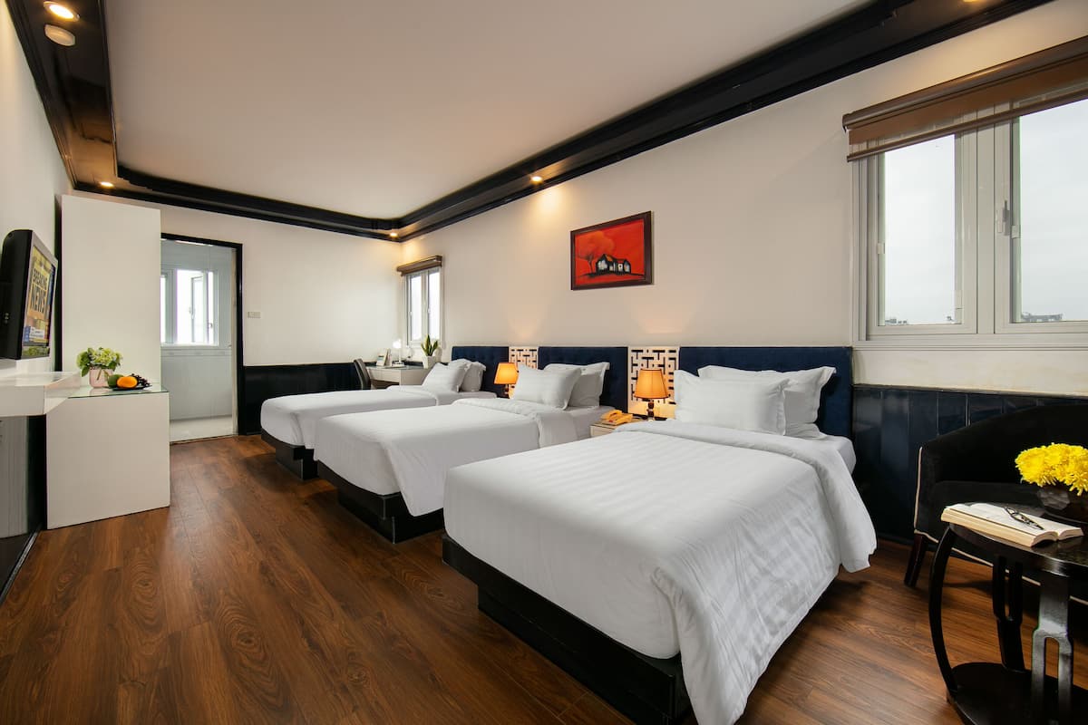 Quarto de hotel com quatro camas individuais, uma escrivaninha com computador, janela com vista e um quadro vermelho na parede.