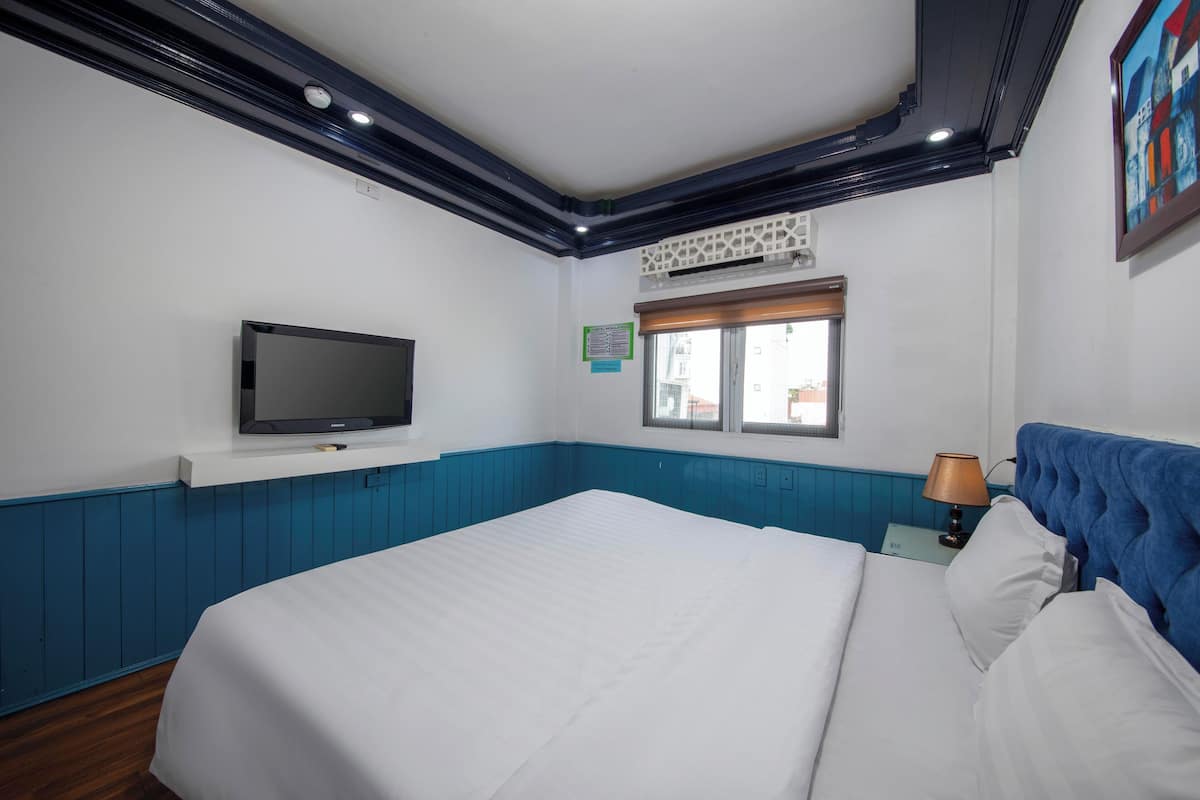 Quarto de hotel com uma cama grande, televisão na parede, janela com persianas e mesa de cabeceira com abajur.