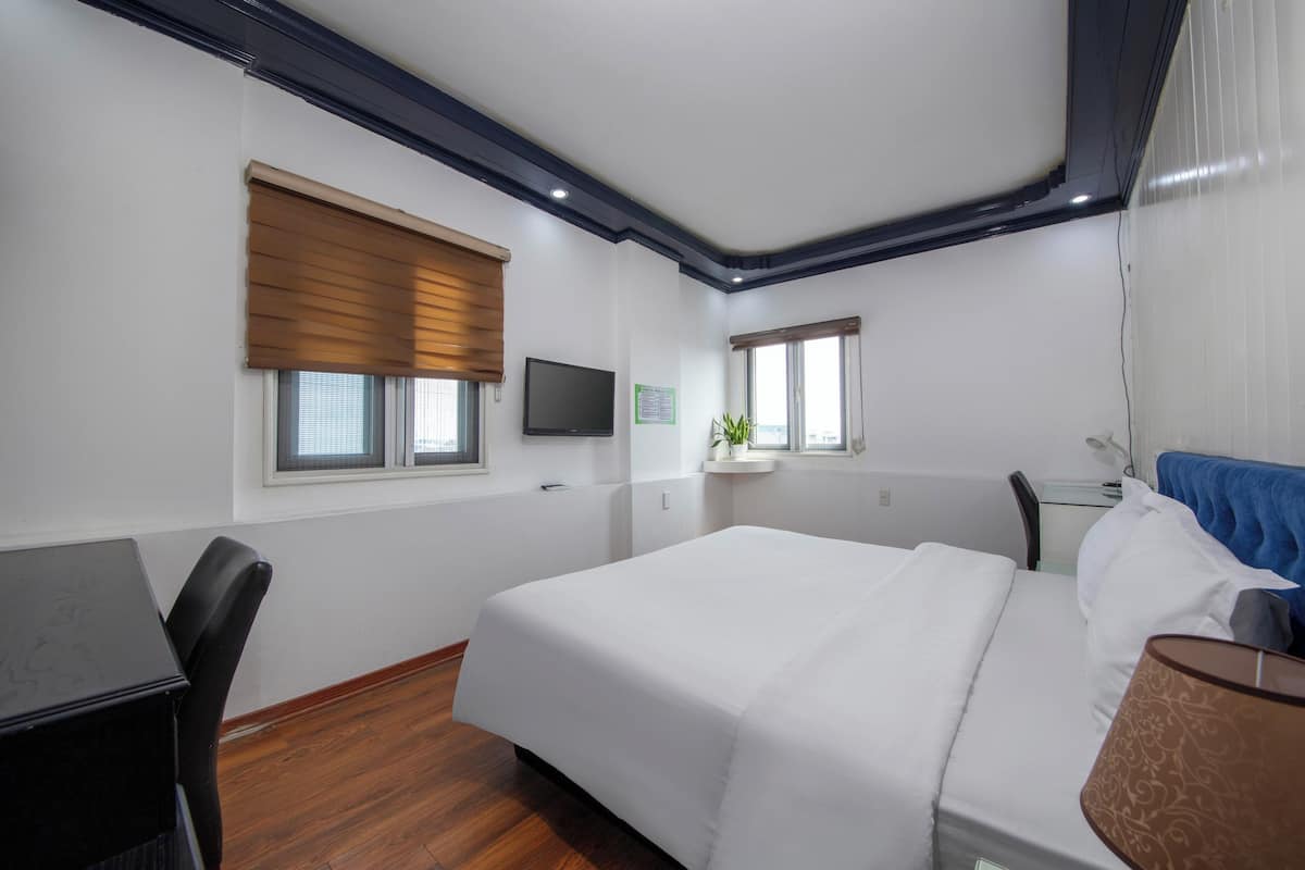Quarto de hotel com uma cama grande, uma escrivaninha, uma cadeira, uma televisão e uma janela com persianas.