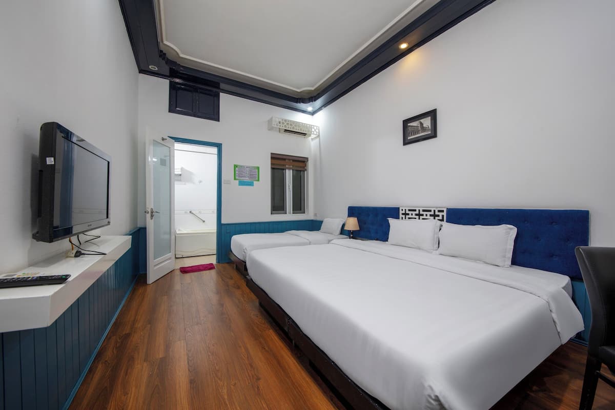 Quarto de hotel com uma cama grande, televisão em móvel, escrivaninha com computador e porta para o banheiro.