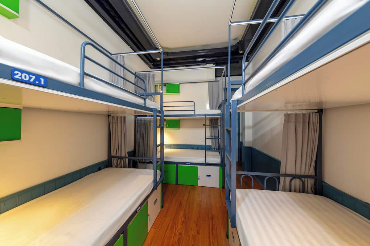 Quarto com beliche de madeira, chão de madeira, beliches verdes e azuis, e um aviso numerado na parede.
