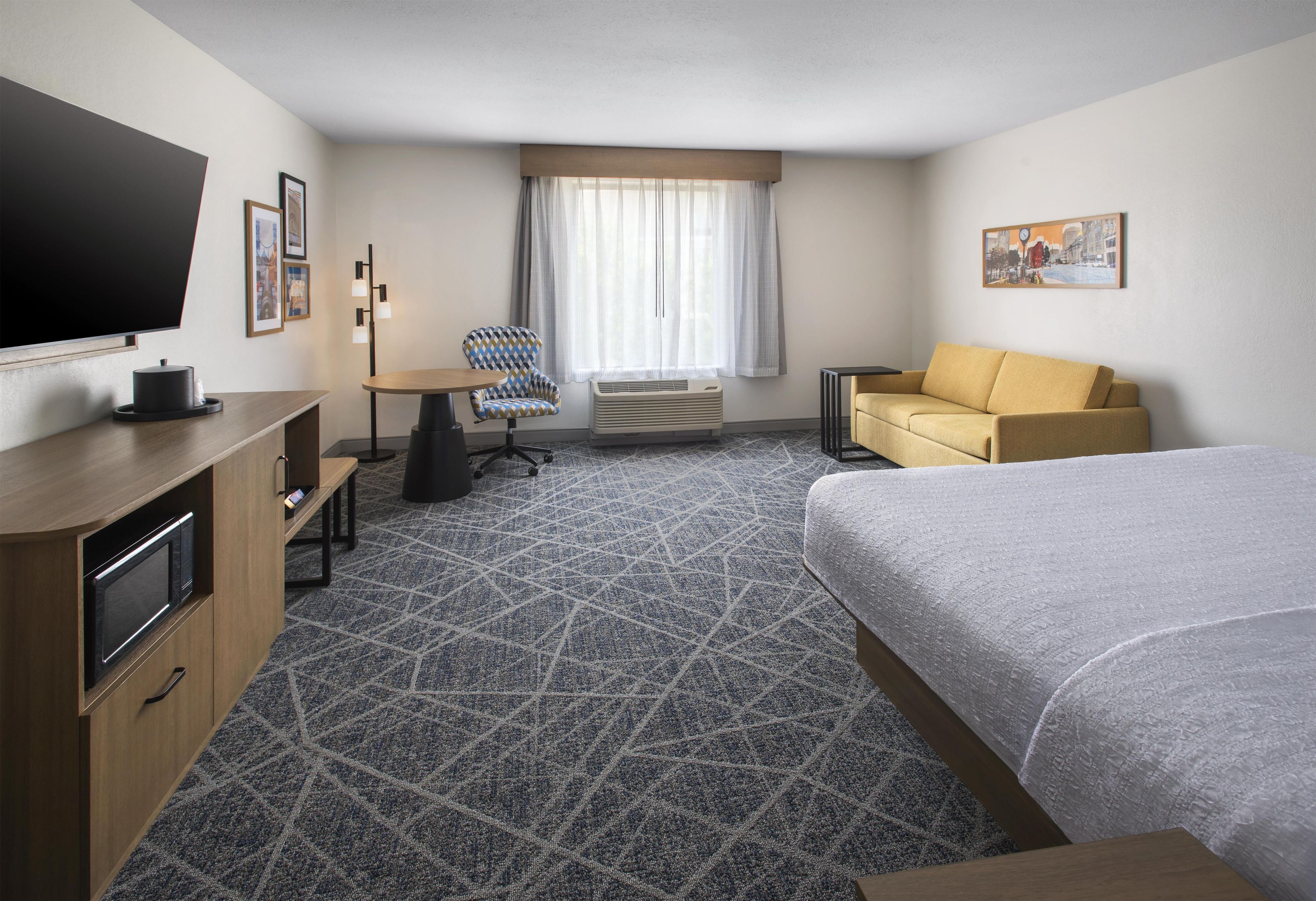 Quarto de hotel com cama, sofá, escrivaninha com cadeira, televisão e janela com cortinas.