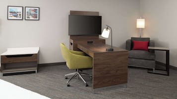 Quarto de hotel com escrivaninha de madeira, uma cadeira verde, um sofá cinzento, uma televisão, mesa de cabeceira com abajur e duas fotografias emolduradas na parede.