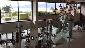 Um hall de hotel moderno com escadaria de vidro, áreas de estar e grandes janelas com vista para um lago e montanhas.