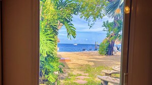 Visão através de uma porta, revelando uma área tropical externa com um caminho que leva a uma praia, palmeiras e um céu claro.