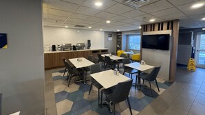 Uma sala de jantar moderna com piso estampado em formato hexagonal, mesas brancas e cadeiras pretas. É visível ao fundo um balcão com uma máquina de café e um quadro de menu.
