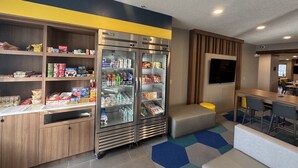 Uma cozinha compacta moderna, com geladeira embutida, prateleiras com petiscos e bebidas, e um espaço de estar com banco e TV fixada na parede.