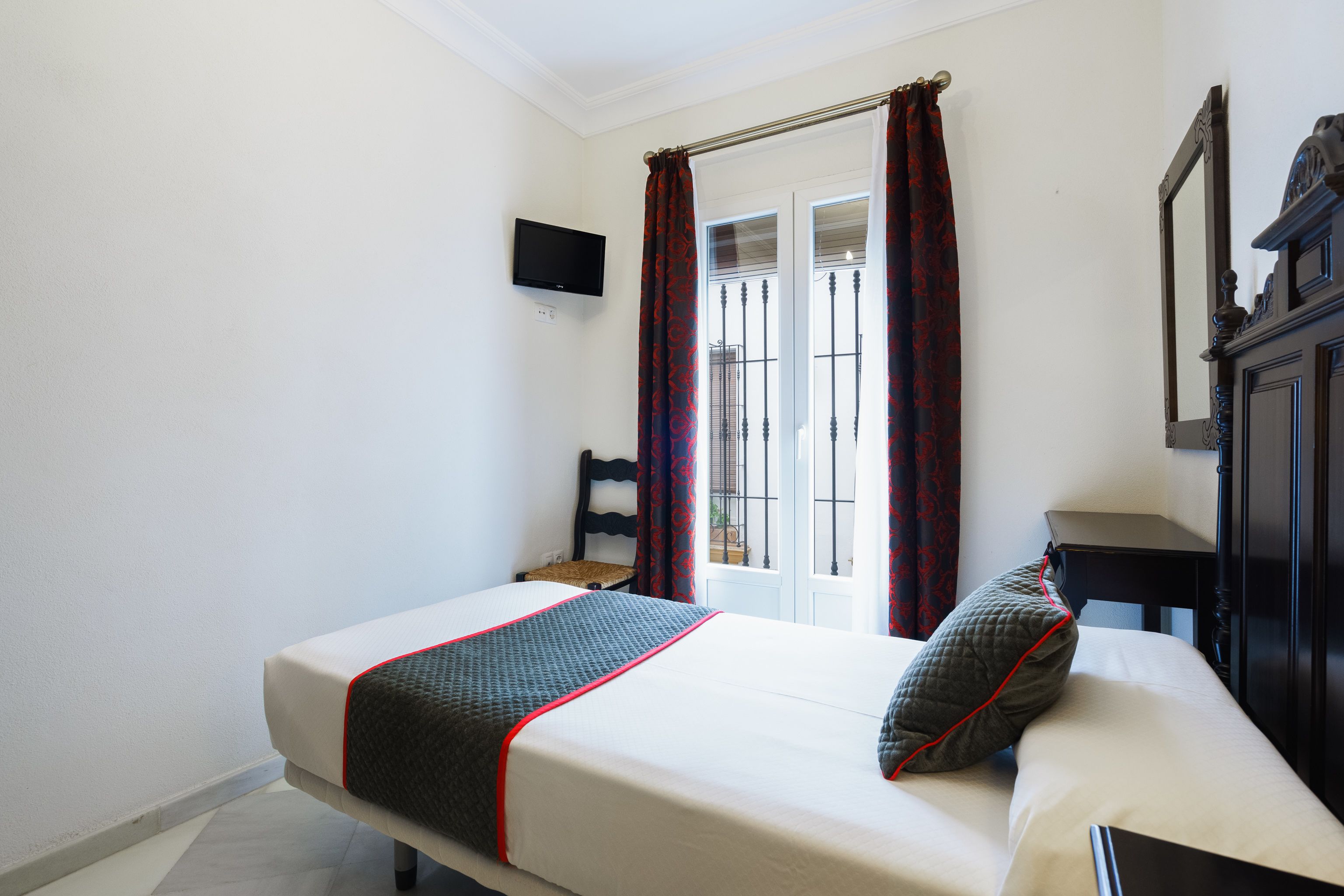 Um quarto com uma cama, uma TV fixada na parede, uma janela com cortinas e uma porta com vista.
