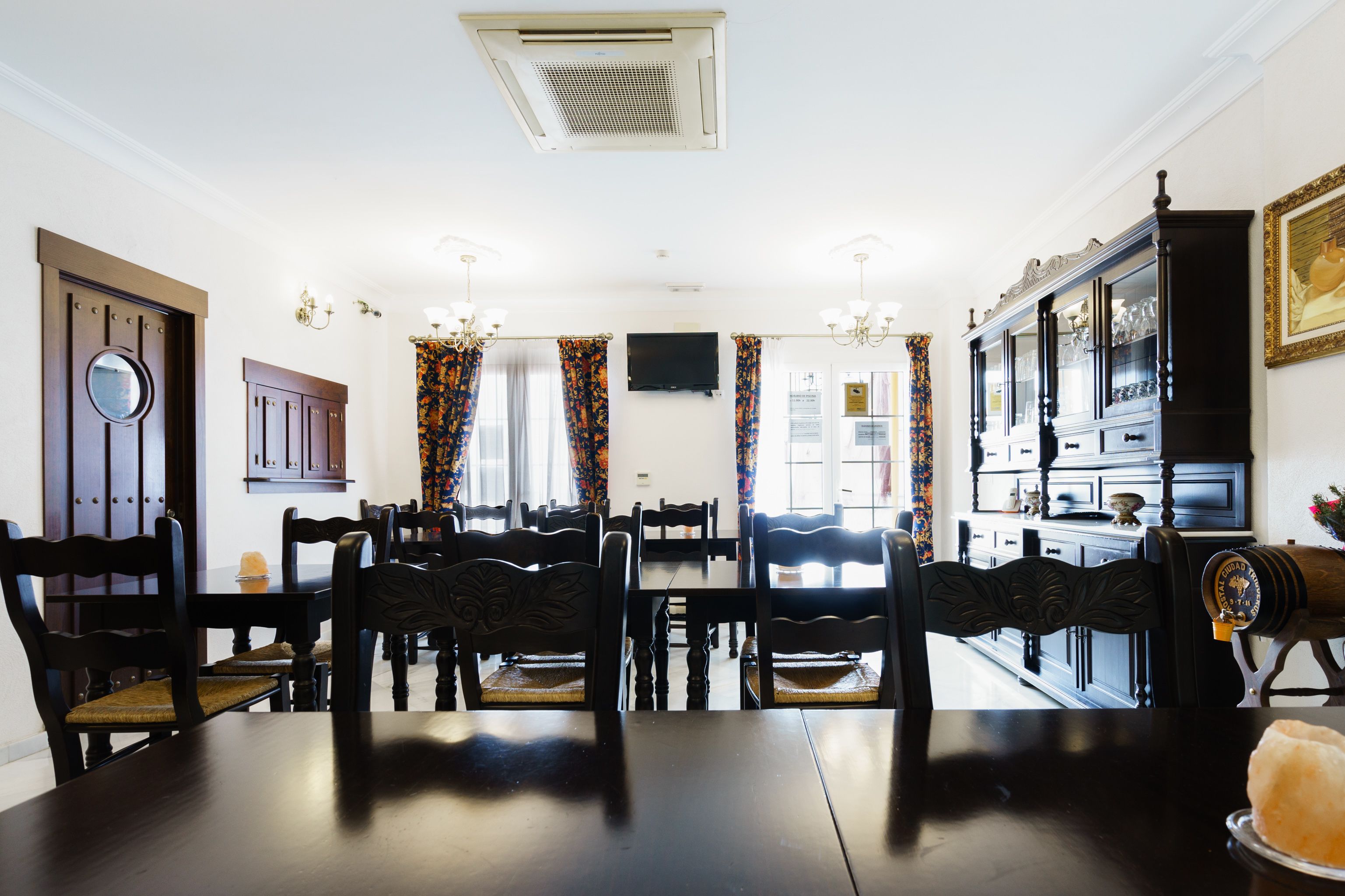 Uma sala de jantar com uma mesa comprida e cadeiras, uma televisão na parede e um armário com portas de vidro.