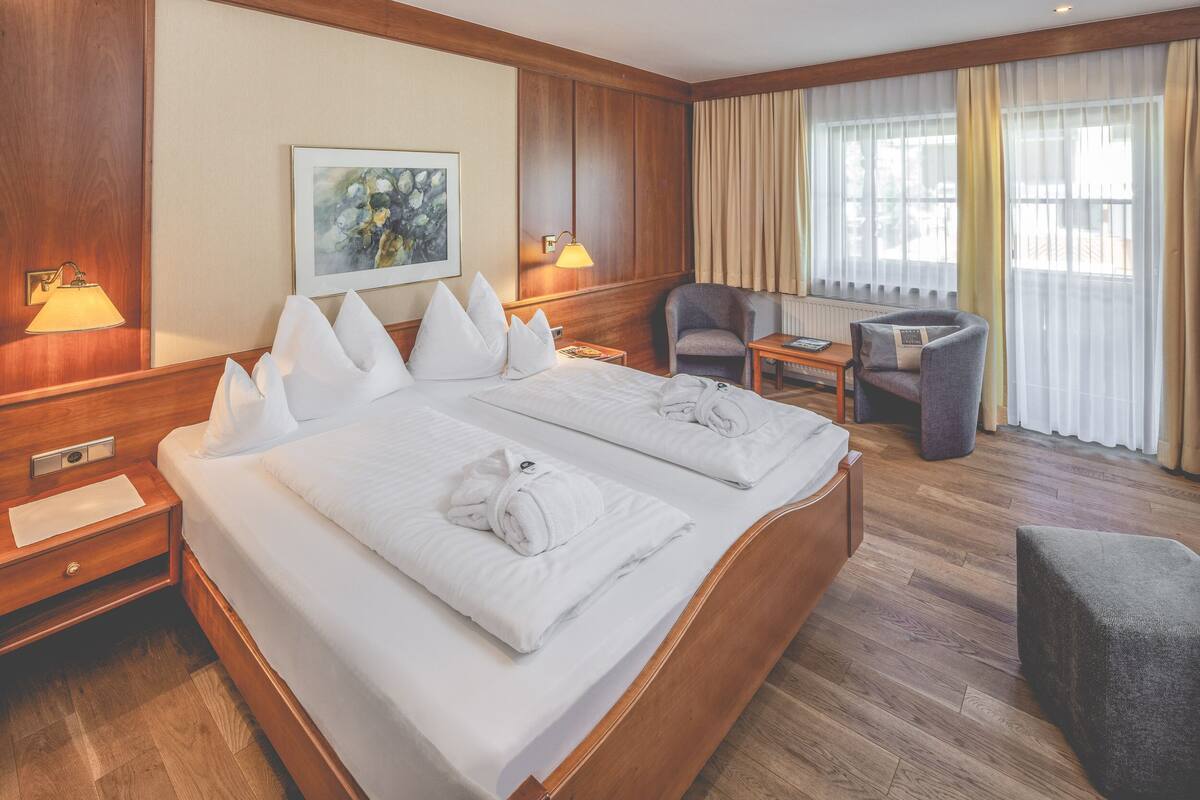 Quarto de hotel com uma cama grande, duas mesas de cabeceira, uma cadeira, uma mesinha e uma janela com cortinas.