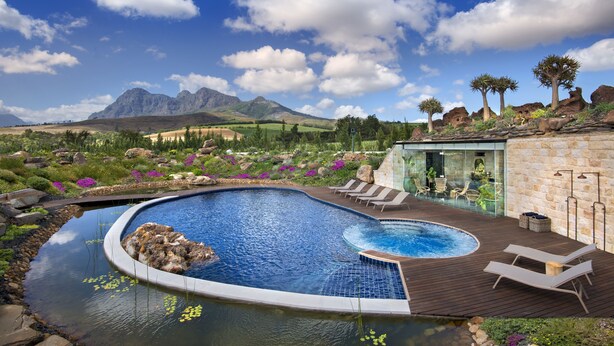 Piscina com deck de madeira e espreguiçadeiras, rodeada por pedras e plantas, com montanhas ao fundo.