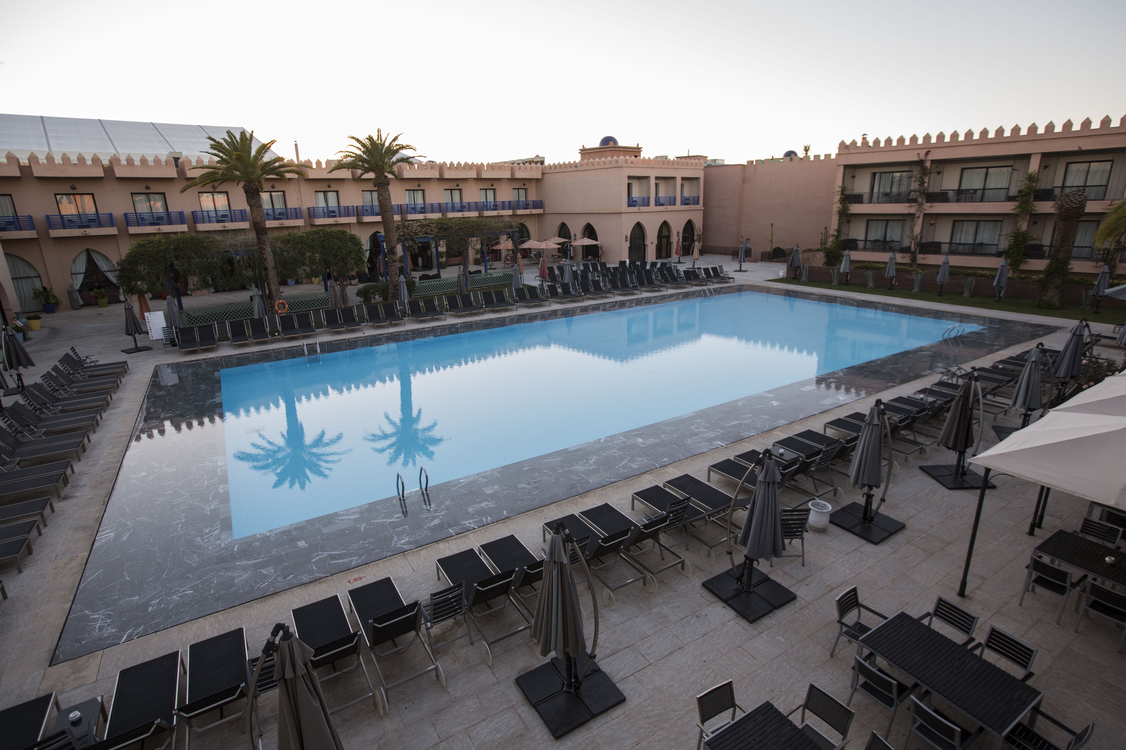 Piscina com cadeiras de descanso e guarda-sóis, cercada por um edifício com varandas e piso de azulejos.