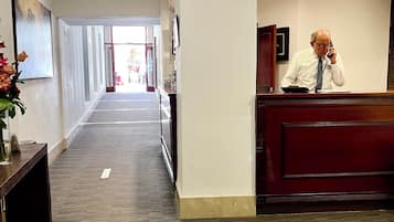 Uma área de recepção de hotel com recepcionista, balcão de atendimento, piso coberto de carpete e um corredor que leva à entrada.