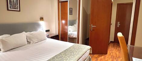 Quarto de hotel com uma cama grande, uma escrivaninha com cadeira, um guarda-roupa e um espelho.