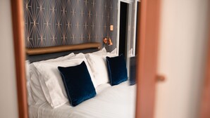 Uma cama bem arrumada com almofadas brancas e azul-escuras, uma cabeceira de madeira e uma parede ao fundo com padrão geométrico.