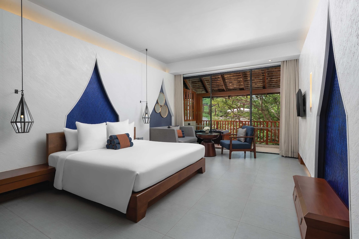 Quarto de hotel com uma cama grande, um sofá, uma mesa de jantar e uma varanda com vista para o verde.