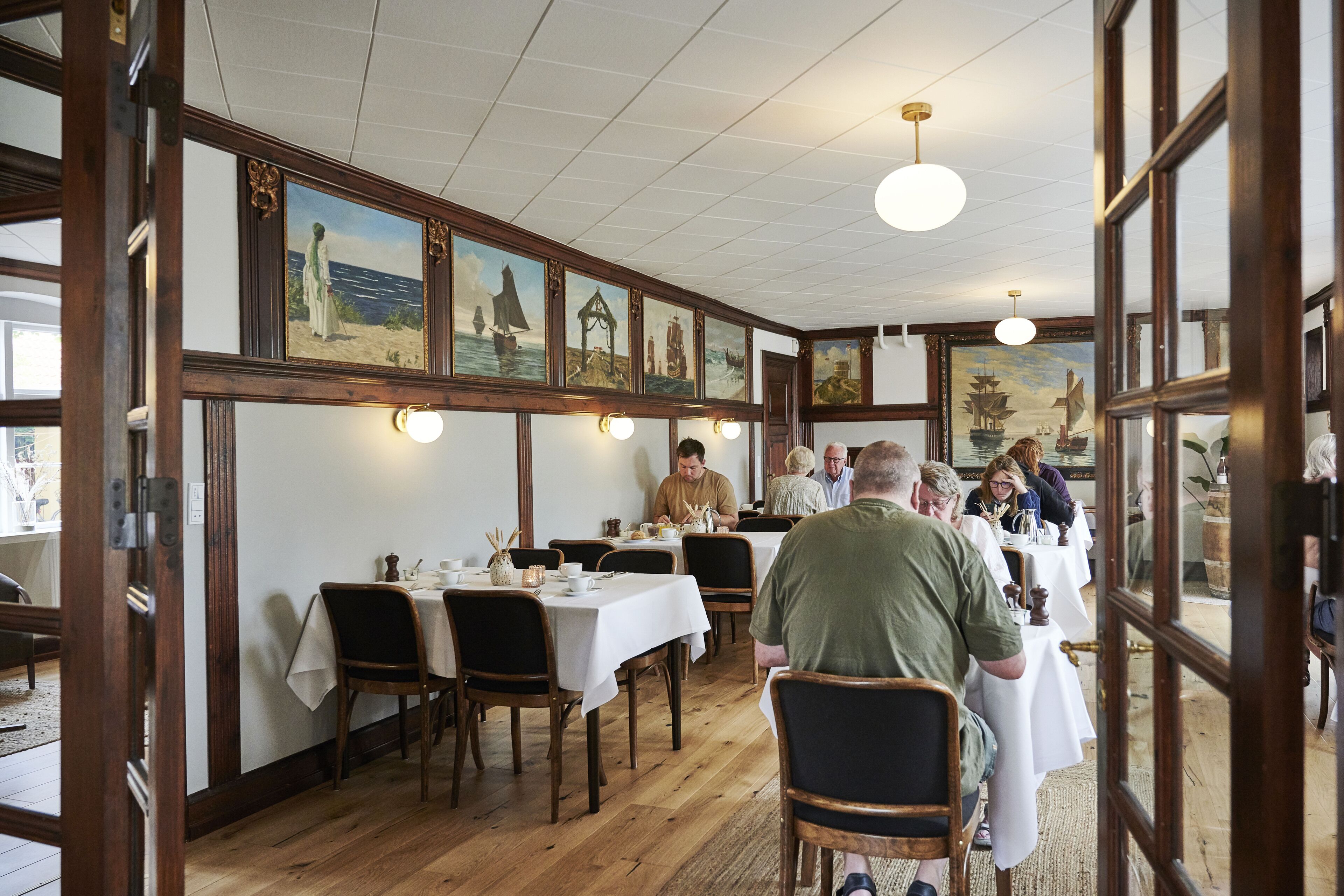 Uma sala de jantar com piso de madeira, mesas e cadeiras brancas. Há pinturas nas paredes retratando cenas marítimas.