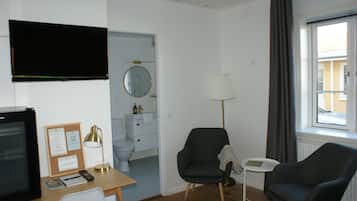 Quarto de hotel com escrivaninha, duas cadeiras, televisão, espelho, luminária, vaso sanitário e janela com cortinas.