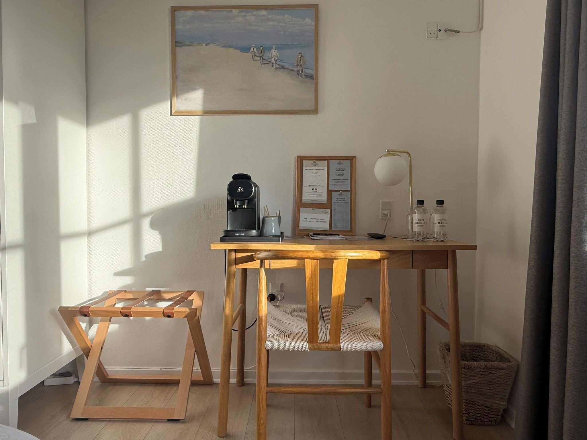 Quarto pequeno e bem organizado, com uma escrivaninha de madeira, uma cadeira, uma cafeteira, um abajur e um quadro na parede.