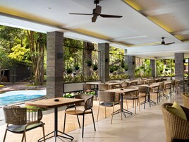 Uma área de refeições ao ar livre com mobiliário de vime, piscina e janelas amplas com vista para um jardim.