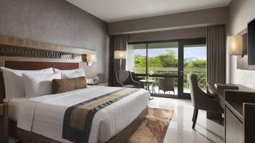 Quarto de hotel moderno com uma cama grande, uma escrivaninha, uma cadeira e uma varanda com vista para o verde.